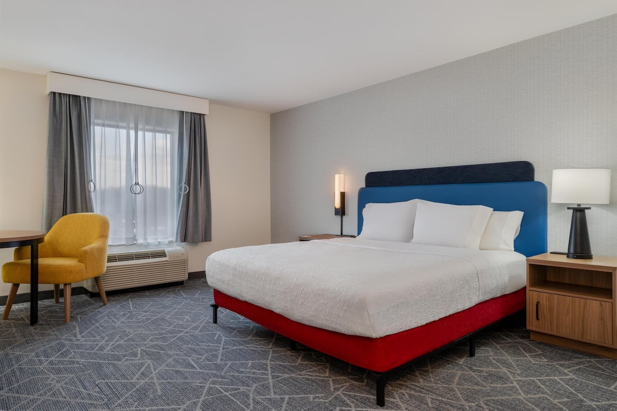 Quarto de hotel com cama, escrivaninha, cadeira, mesa de cabeceira, luminária e janela com cortinas.