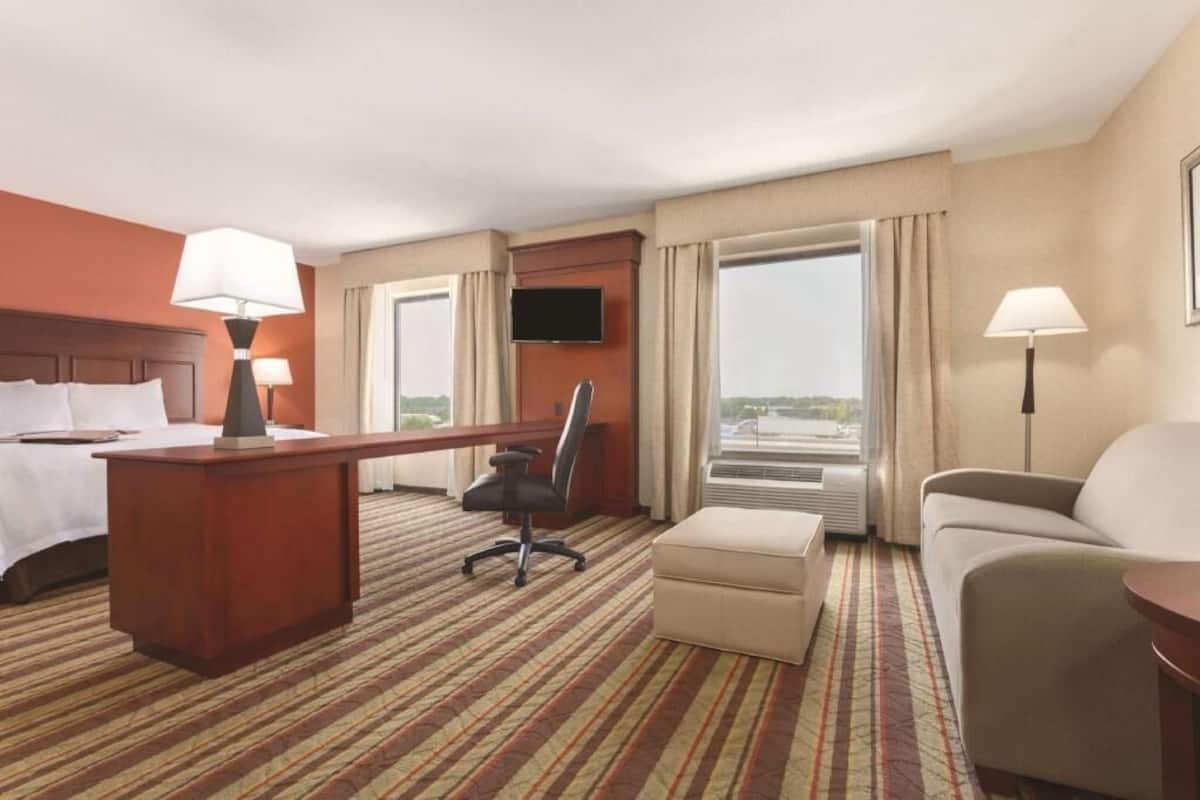 Quarto de hotel com cama, escrivaninha, cadeira de escritório, sofá e televisão na parede.