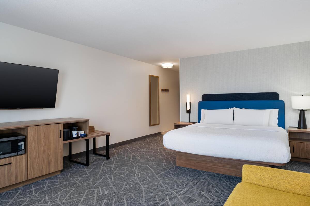 Quarto de hotel com cama, televisão, secretária com micro-ondas e uma pequena mesa.