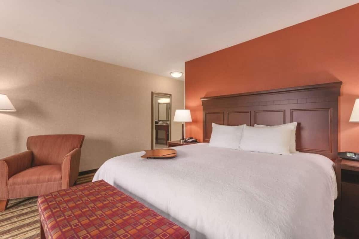 Quarto de hotel com cama, poltrona, banquinho, mesa de cabeceira e luminária.