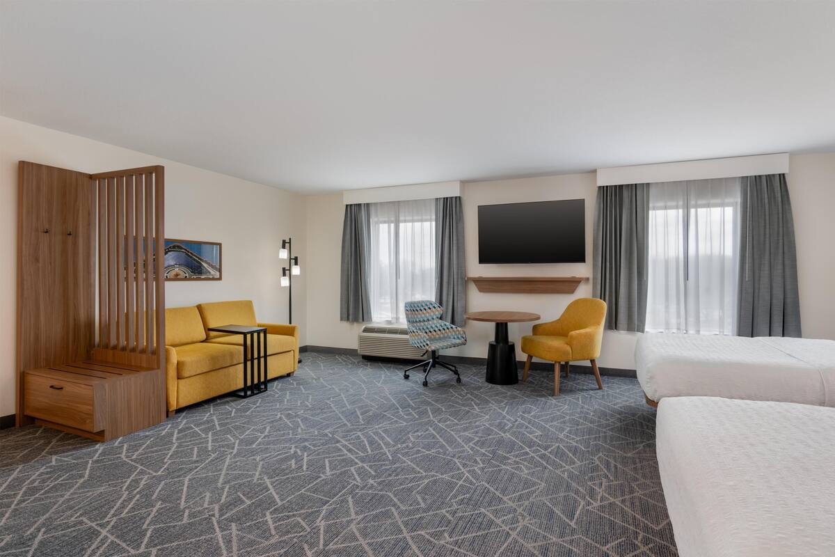 Quarto de hotel com sofá, uma cadeira, mesinha, televisão e cama.