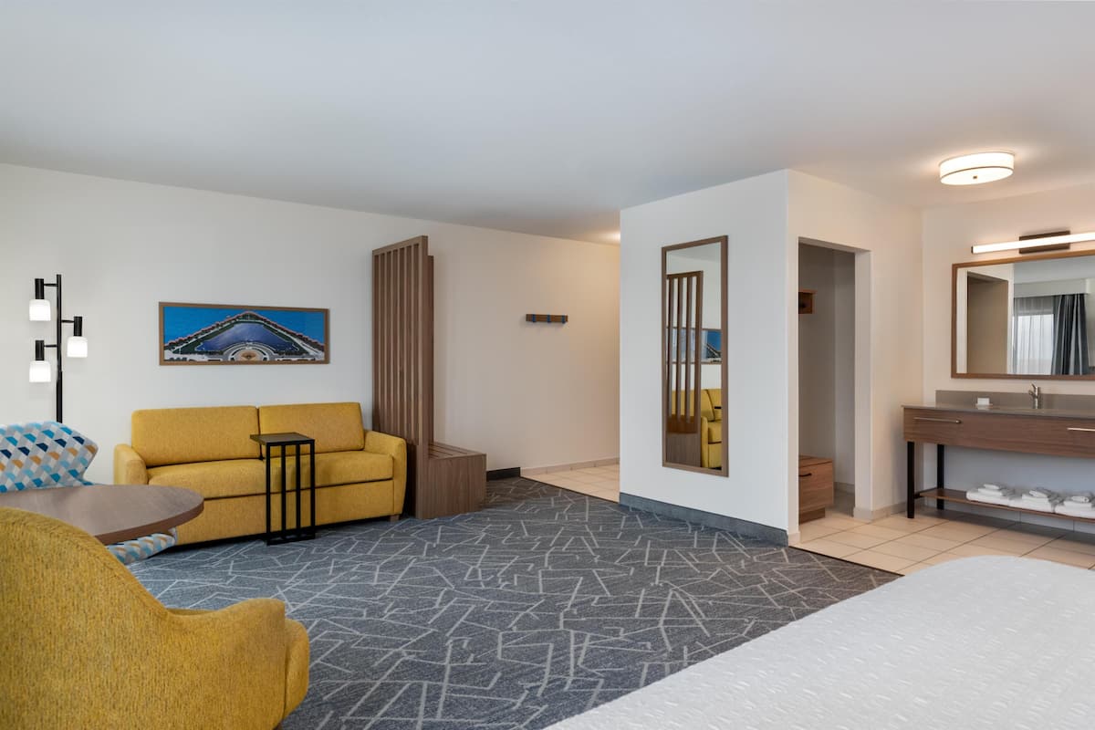 Quarto de hotel com cama, sofá, mesinha, espelho, banheiro com pia e porta-toalhas, e um quadro na parede.