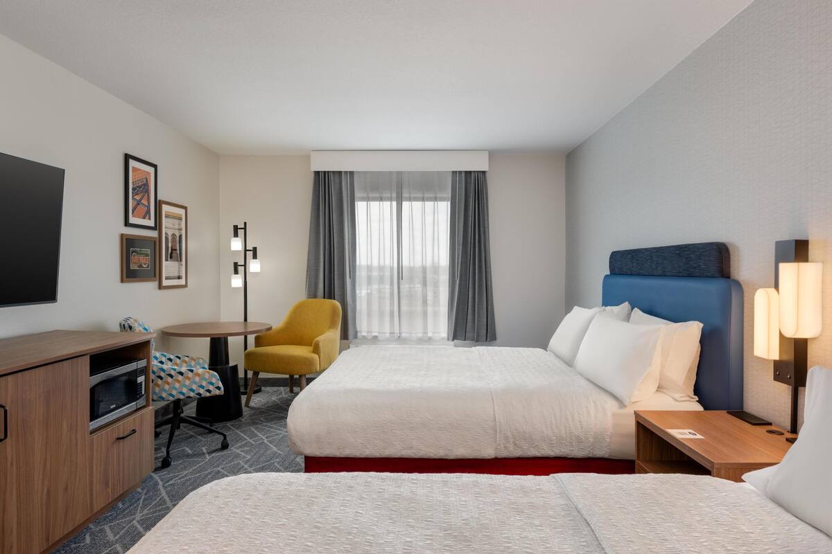Quarto de hotel com duas camas, uma escrivaninha, uma cadeira, uma televisão e uma janela com cortinas.