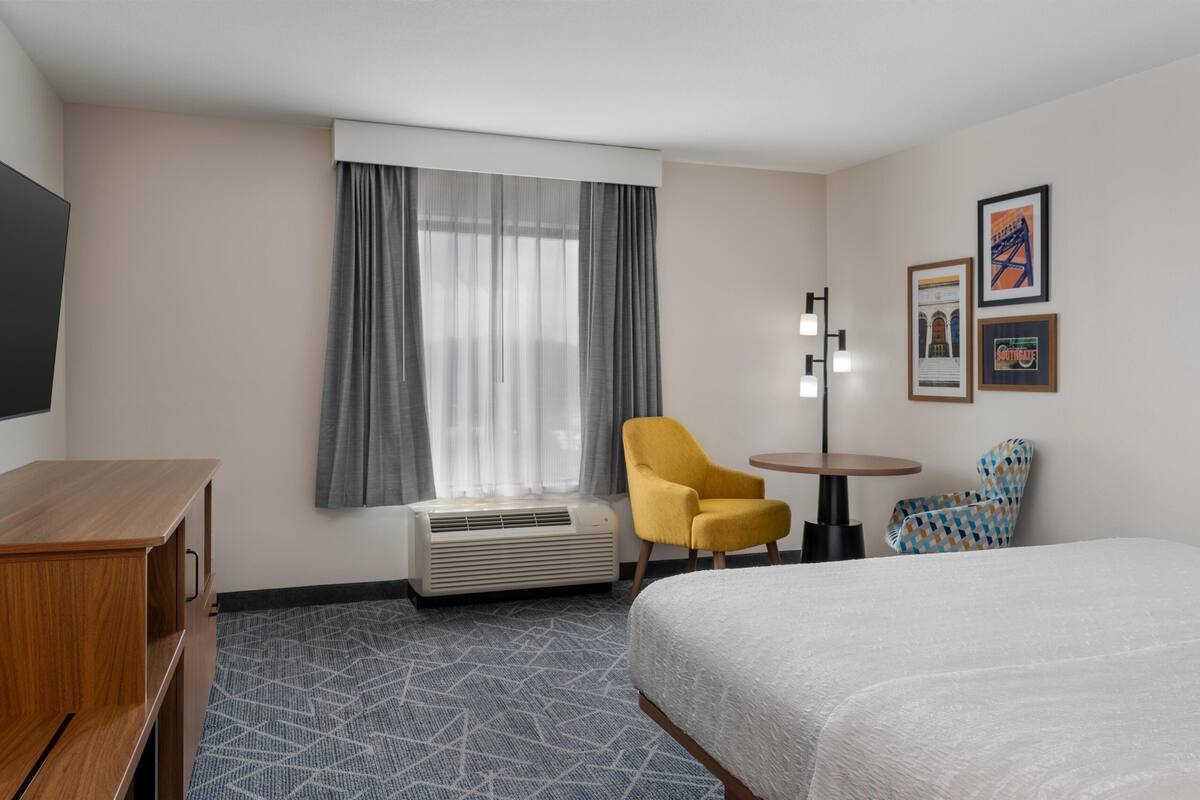 Quarto de hotel com cama, escrivaninha, cadeira, mesa, ar condicionado e janela com cortinas.