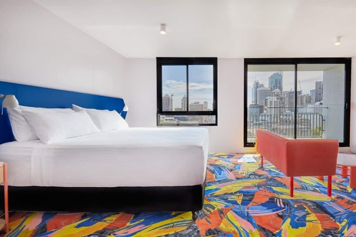 Quarto de hotel com uma cama grande, vista da cidade por uma porta deslizante, piso com padrão abstrato e colorido, e uma cadeira vermelha.