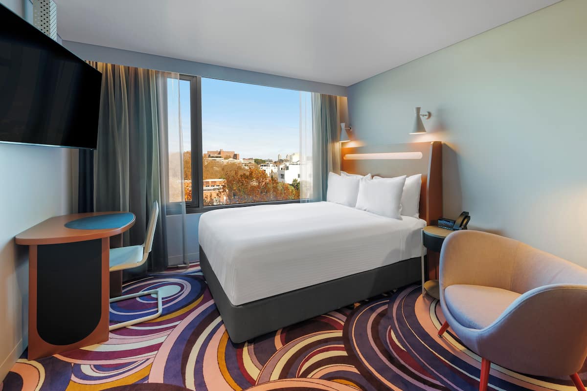 Quarto de hotel com uma cama grande, uma mesa, uma cadeira, uma televisão e vista para a cidade.