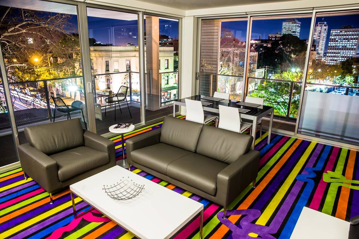 Uma sala de estar moderna com um sofá, cadeiras e uma mesa de centro. Grandes janelas oferecem vista para a cidade.