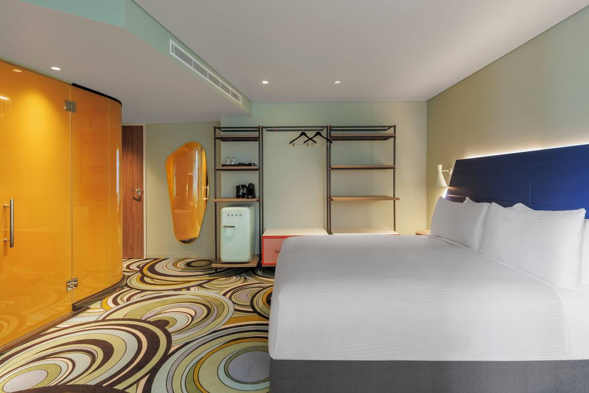 Quarto de hotel com cama grande, guarda-roupa, minibar e prateleira fixada na parede.