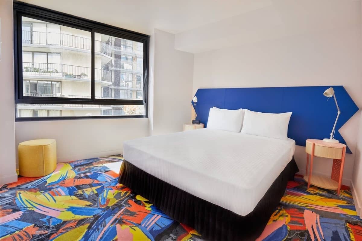 Quarto de hotel com uma cama grande, janela com vista para edifícios, tapete abstrato colorido e um banquinho redondo pequeno.