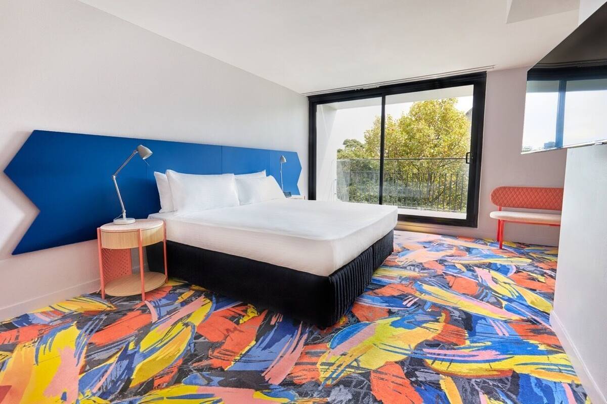 Quarto de hotel moderno com cama grande, mesa de cabeceira, design de chão abstrato e colorido, janela ampla com vista para árvores e uma cadeira vermelha.