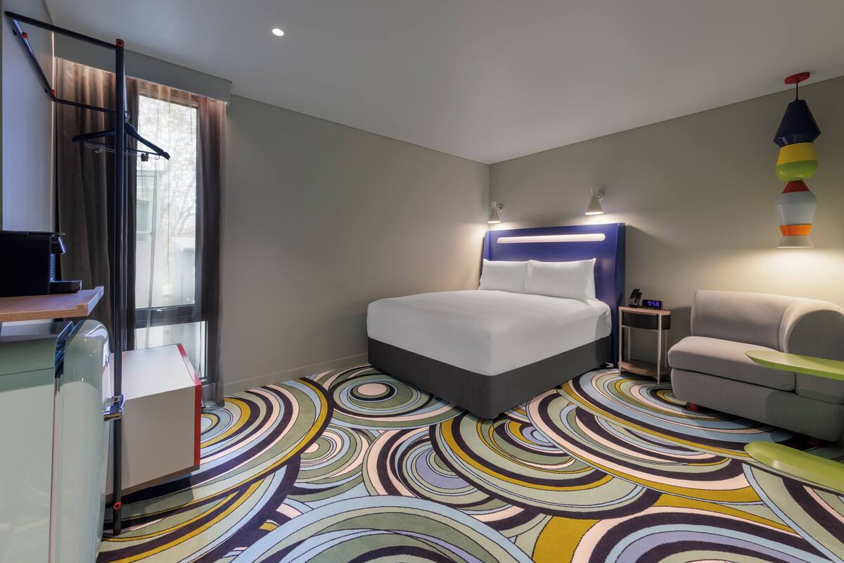 Quarto de hotel com cama, sofá, escrivaninha e um tapete circular colorido.