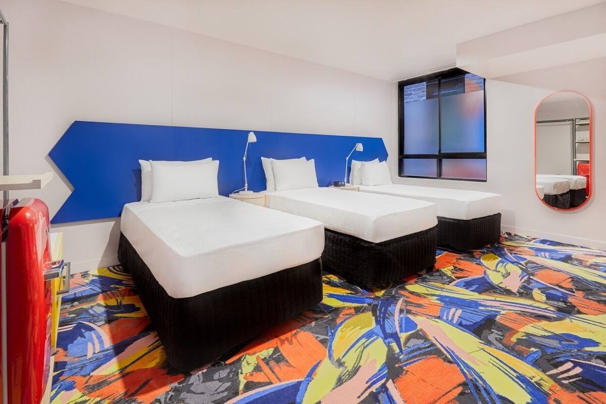Quarto de hotel com duas camas, um espelho grande e um tapete colorido com padrão abstrato.