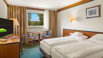 Quarto de hotel com duas camas, uma escrivaninha com televisão, uma cadeira, uma mesinha e uma janela com cortinas.