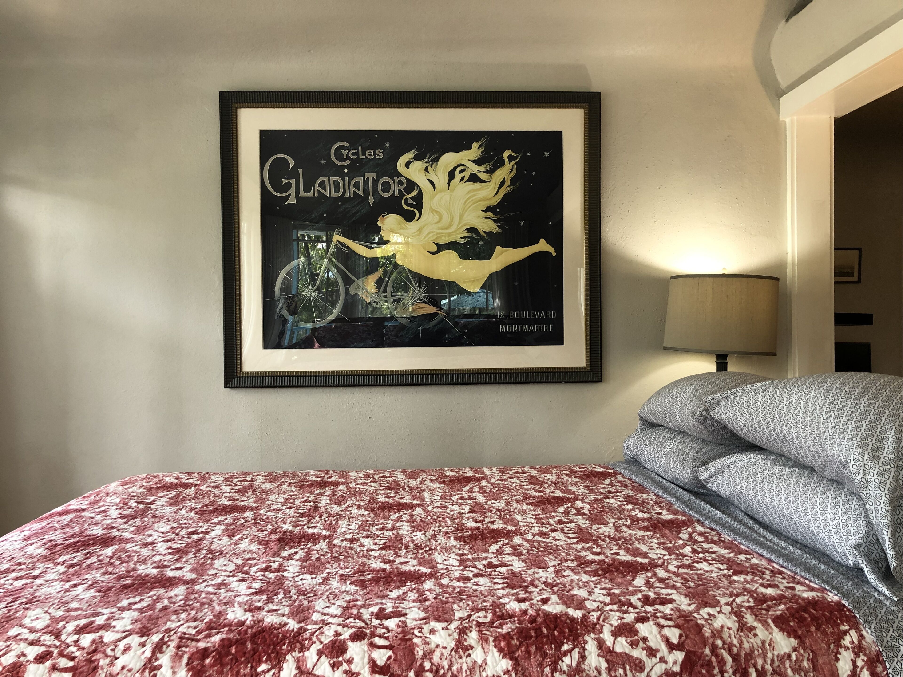 Uma obra de arte emoldurada retrata uma mulher andando de bicicleta em uma parede, uma cama com roupa de cama estampada e um abajur em uma mesa de cabeceira.