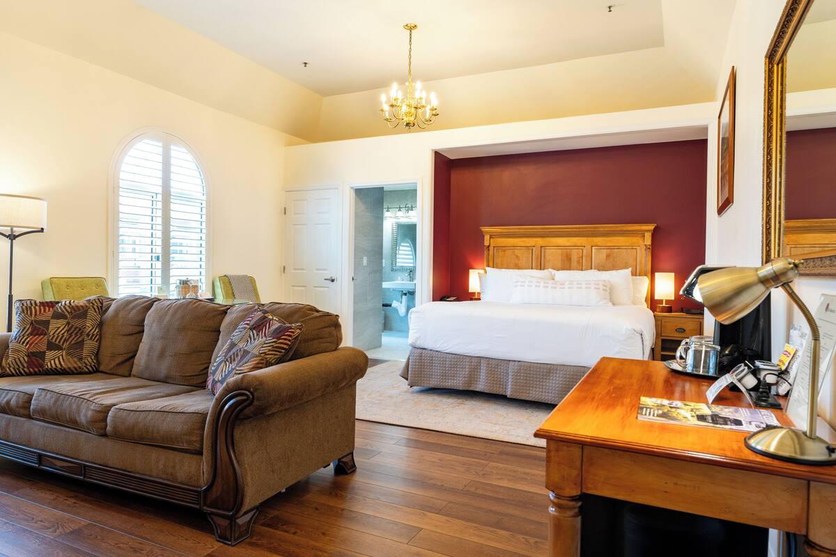 Quarto de hotel com piso de madeira, sofá marrom, cama com lençóis brancos, mesa de cabeceira de madeira, escrivaninha com luminária e um espelho.