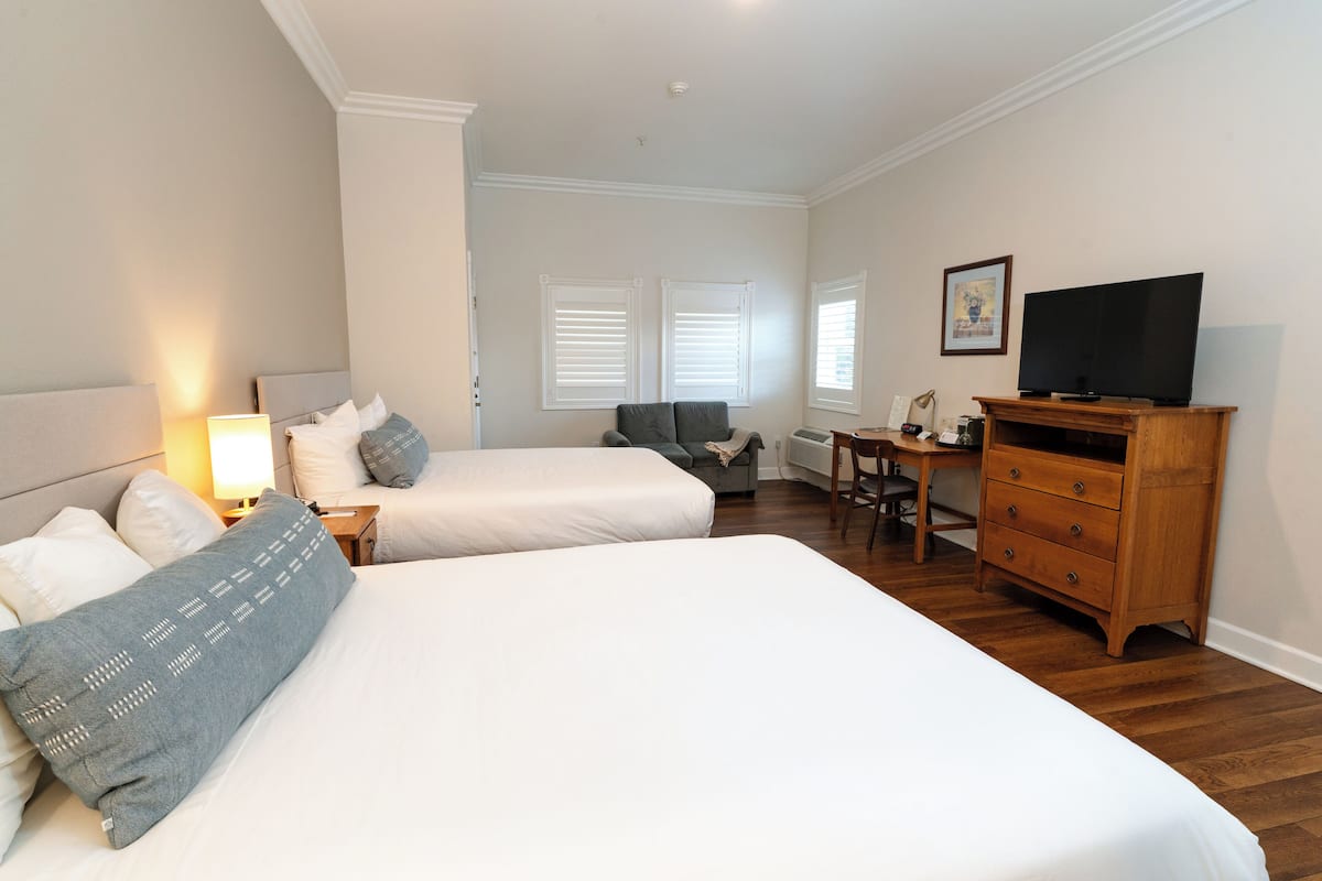 Quarto de hotel com duas camas, uma cômoda de madeira, uma escrivaninha e uma televisão.