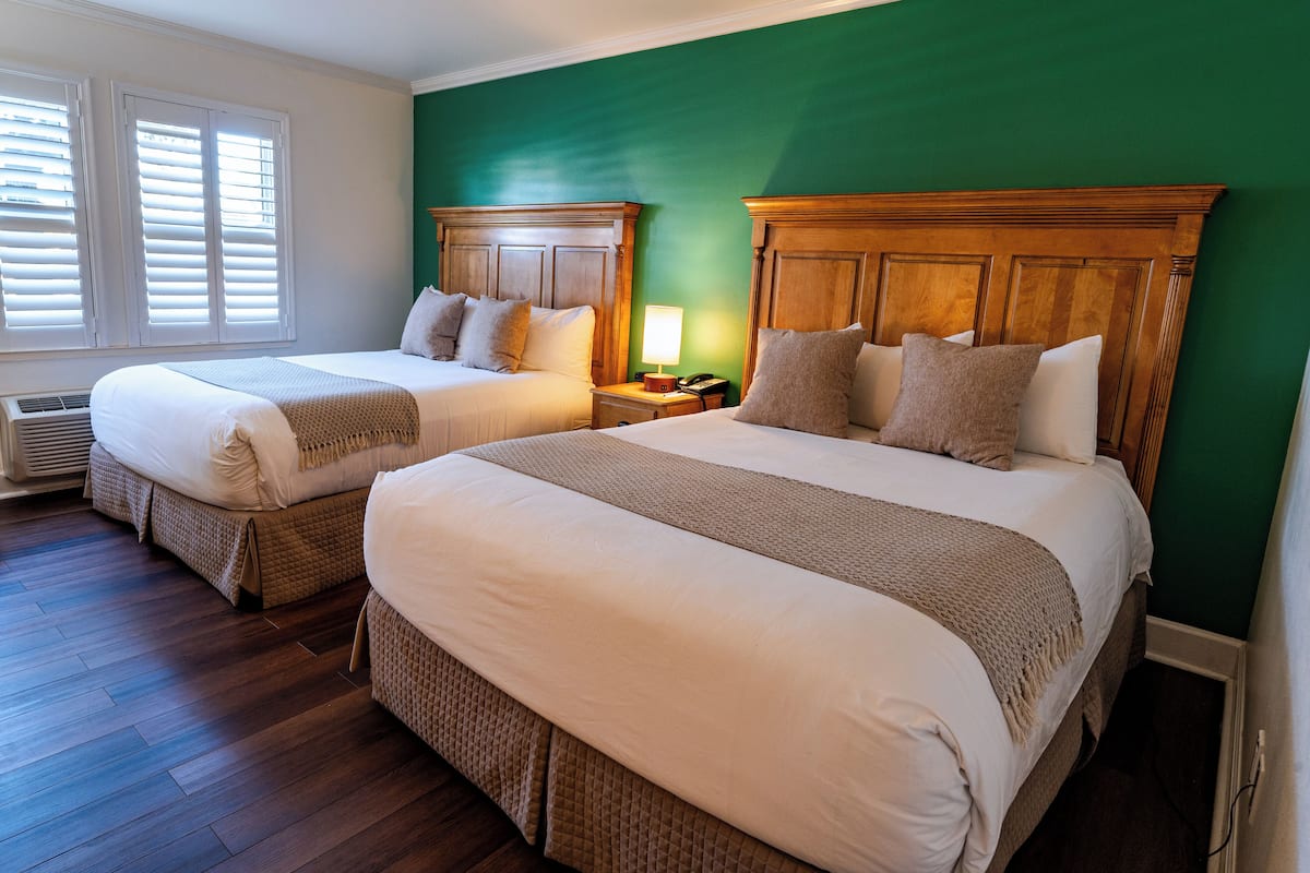 Quarto de hotel com duas camas, cabeceiras de madeira, parede verde decorativa e janela com persianas.