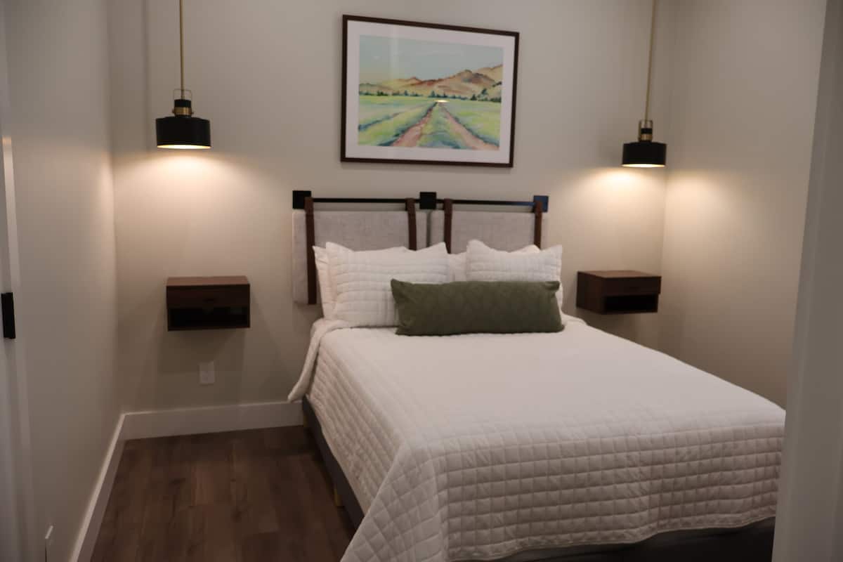 Cama bem arrumada com roupa de cama branca e almofada verde, com duas mesas de cabeceira laterais e luminárias fixadas na parede.