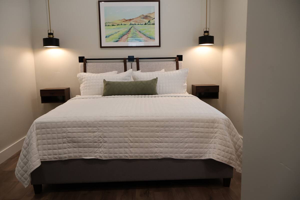 Cama bem arrumada com colcha acolchoada branca e dois travesseiros, com mesas de cabeceira laterais com abajures pretos.