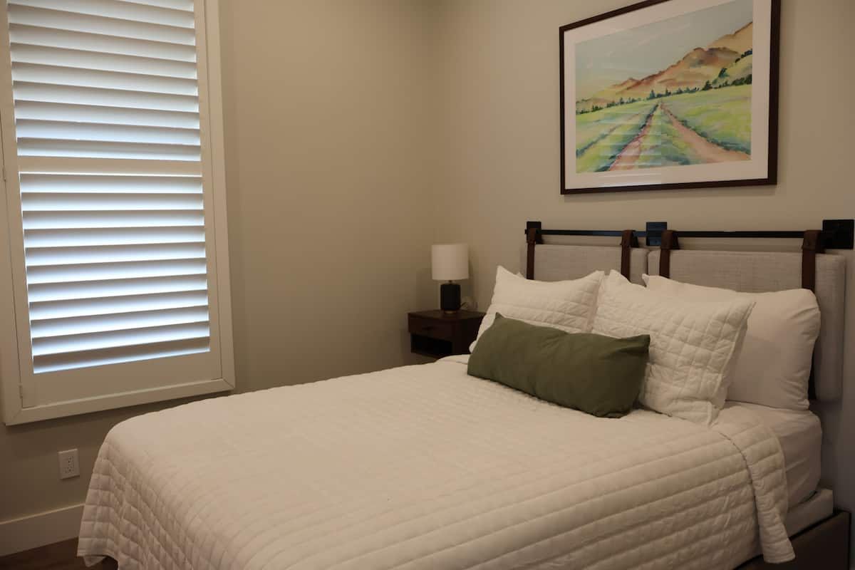 Uma cama bem arrumada com um edredom branco e uma almofada verde, uma mesa de cabeceira com abajur e um quadro na parede.
