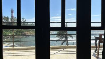Vista de dentro de um edifício para uma área costeira, com um farol, palmeiras e um corpo d'água.