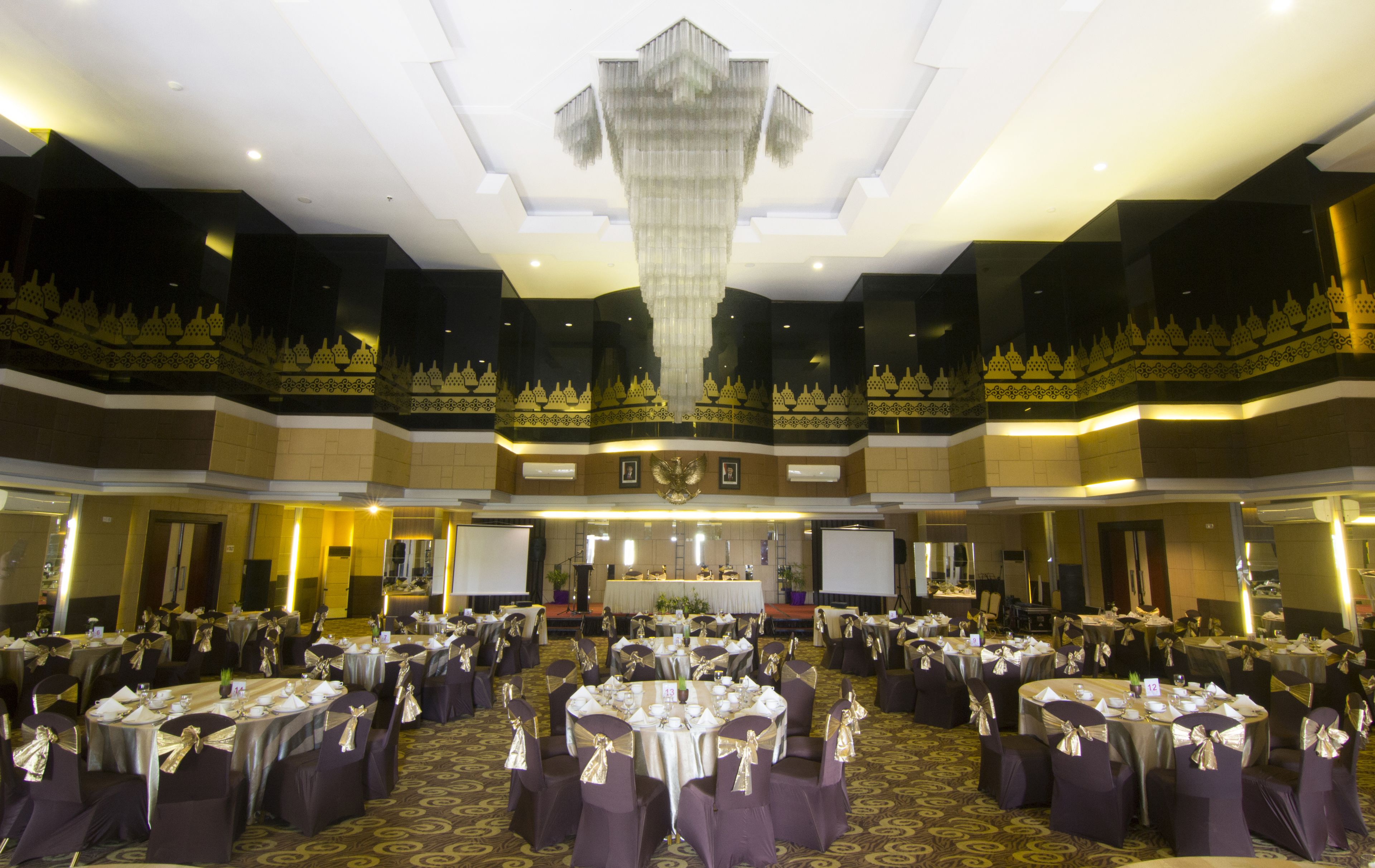 Uma sala de banquetes com mesas redondas preparadas para refeições, um lustre grande e padrões decorativos nas paredes.