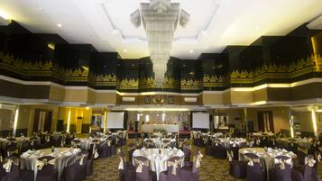 Uma sala de banquetes com mesas redondas preparadas para refeições, um lustre grande e padrões decorativos nas paredes.