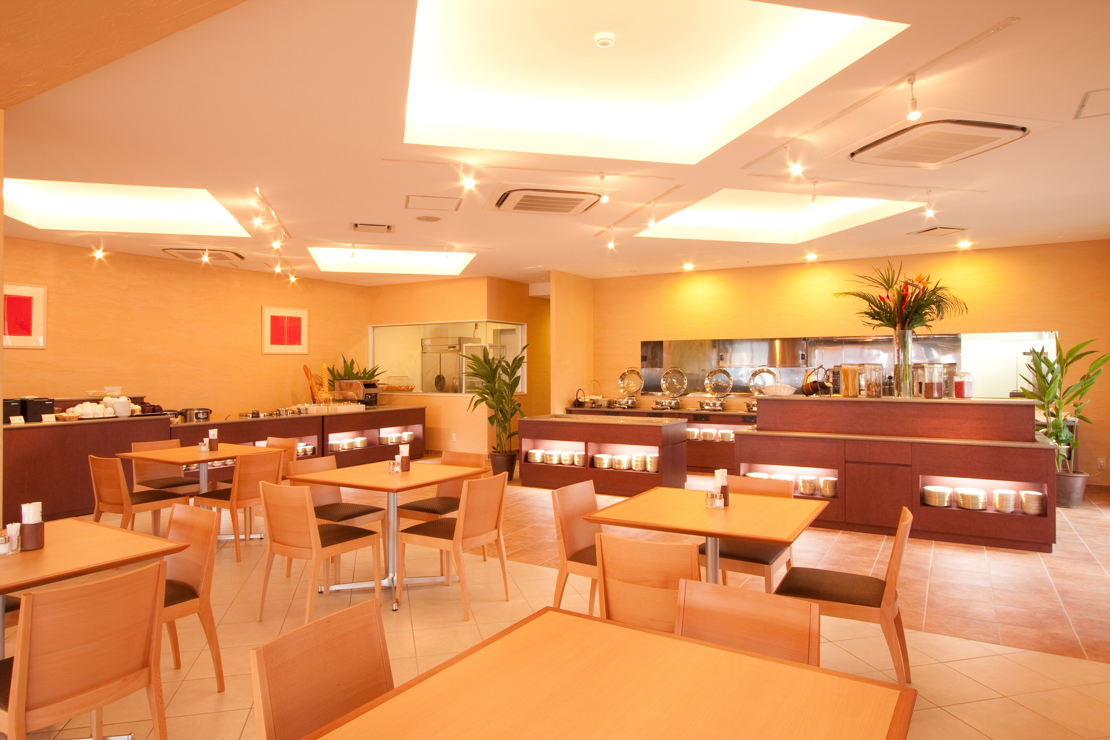 Um restaurante espaçoso com várias mesas e cadeiras, um balcão exibindo diversos alimentos e uma área de cozinha ao fundo.