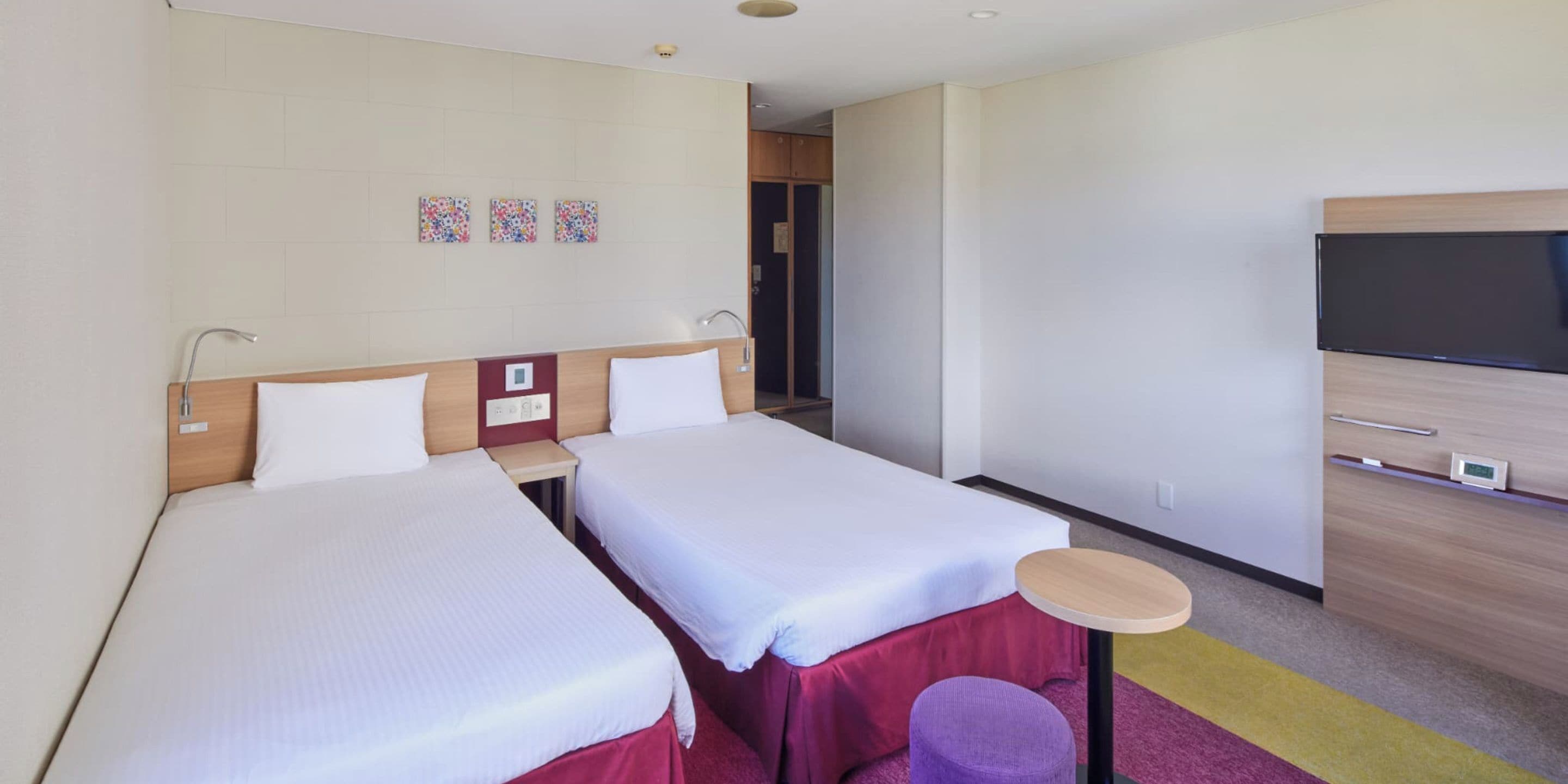 Quarto de hotel com duas camas, uma televisão, uma mesa pequena e um banheiro visível ao fundo.