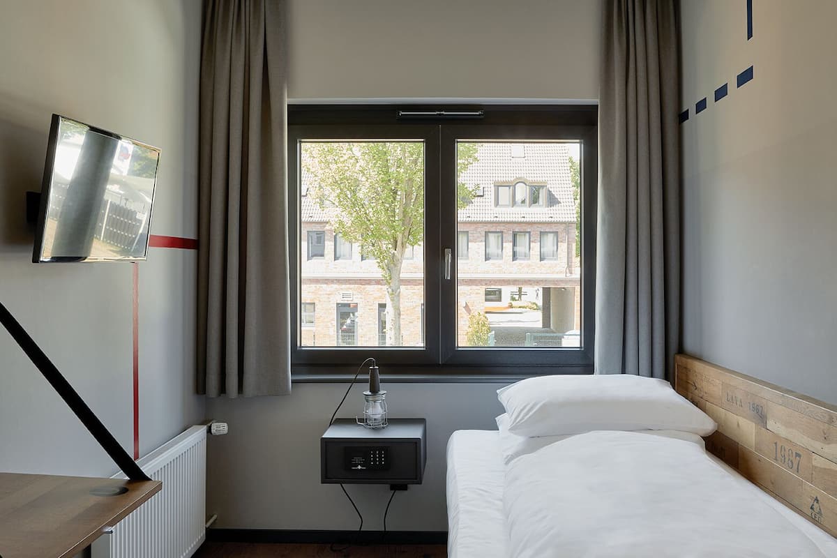 Quarto de hotel com uma cama, televisão fixada na parede, janela com cortinas e radiador.