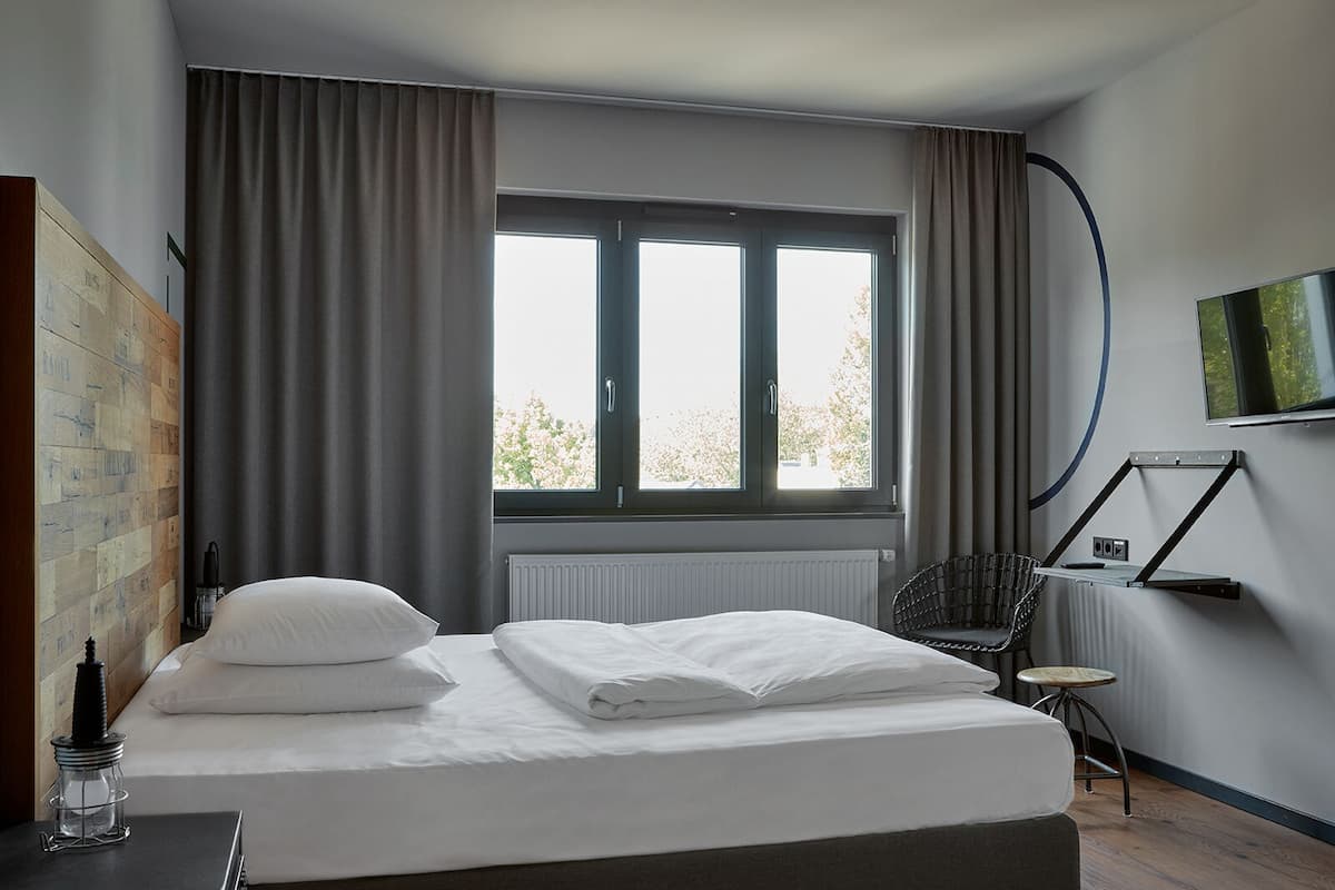 Quarto de hotel moderno com uma cama grande, vista para árvores pela janela, uma cadeira e uma televisão na parede.