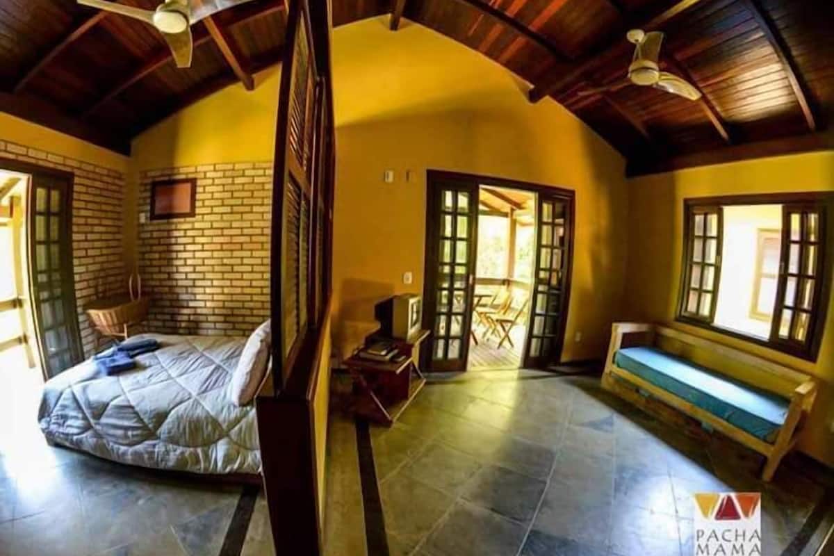 Quarto espaçoso com cama, escrivaninha e um banco. Duas portas dão para área externa.