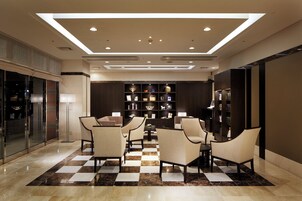 Um hall de hotel com piso xadrez, uma área de estar central com cadeiras, uma estante de livros e um design de teto moderno.