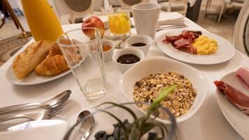 Mesa de café da manhã com croissants, ovos mexidos, bacon e uma tigela de granola.