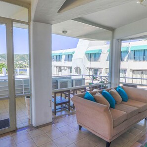 Uma varanda com um sofá bege, uma mesa e cadeiras. Janelas amplas oferecem vista para edifícios e um corpo de água.