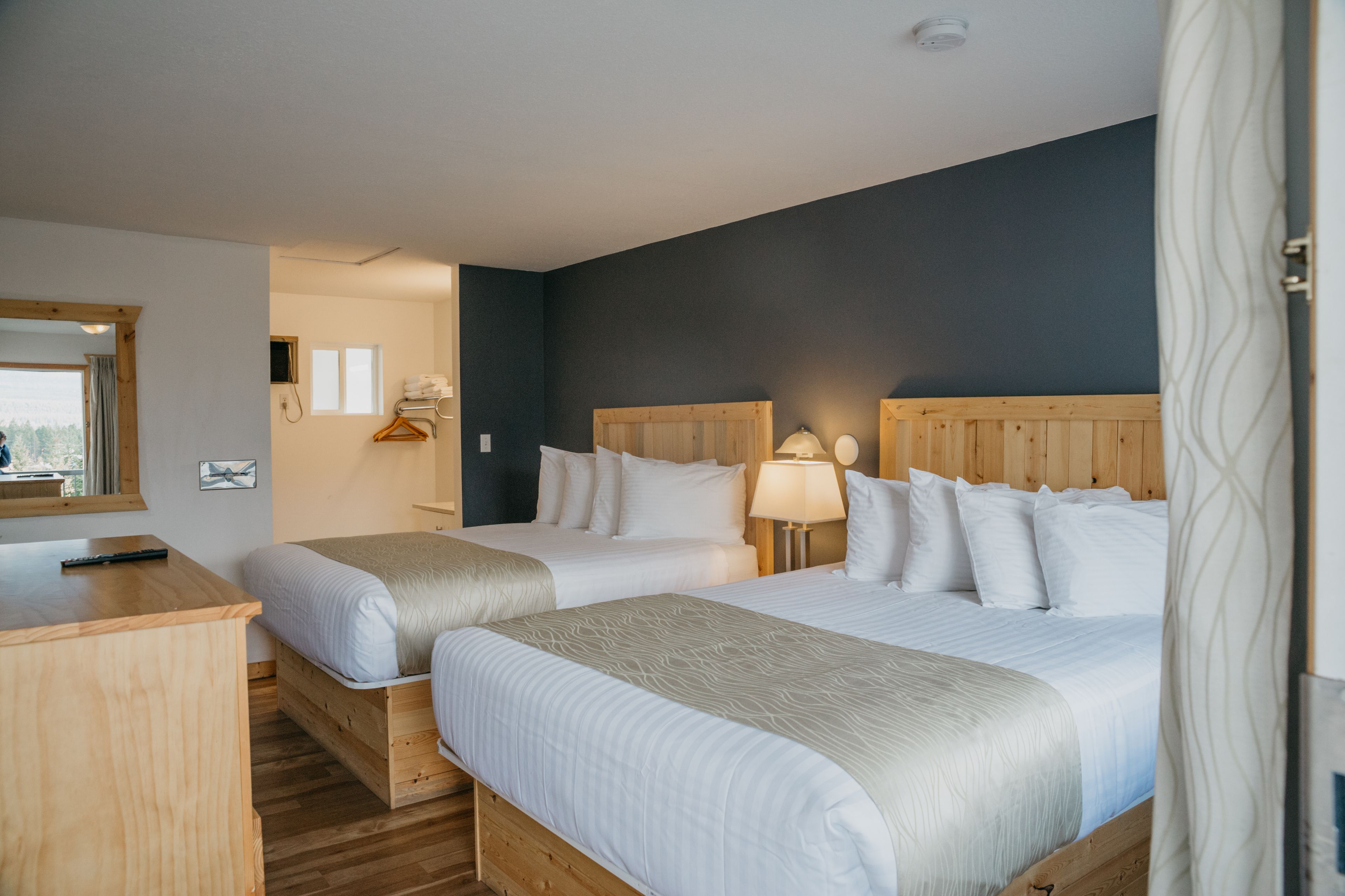 Quarto de hotel com duas camas, uma escrivaninha de madeira, um espelho, um abajur e vista para o exterior.