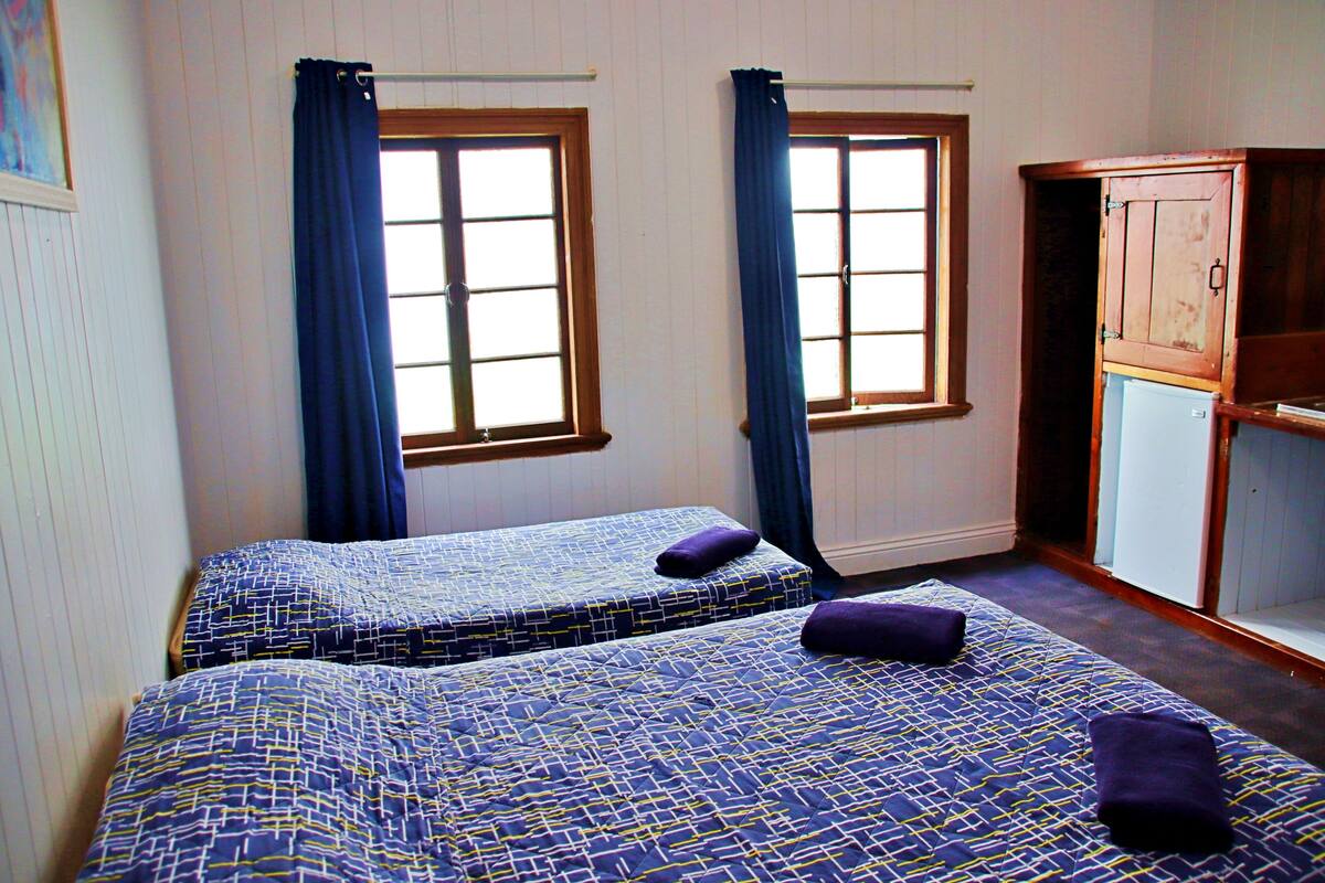 Quarto com duas camas, um armário de madeira e duas janelas com cortinas.