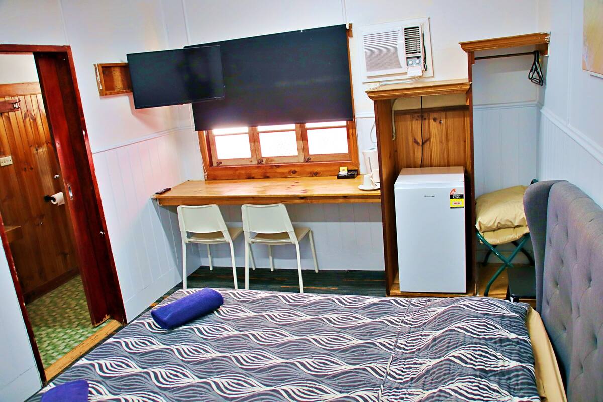 Quarto compacto com cama, escrivaninha com duas cadeiras, pequena geladeira e televisão na parede.
