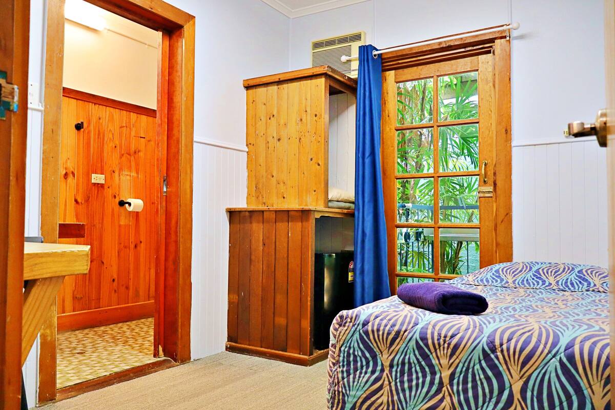 Quarto com cama, guarda-roupa de madeira, pequena cozinha e janela com vista para área verde.