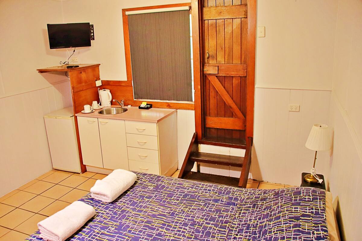Quarto com cama, uma pequena escrivaninha com pia, televisão e janela com porta de madeira.