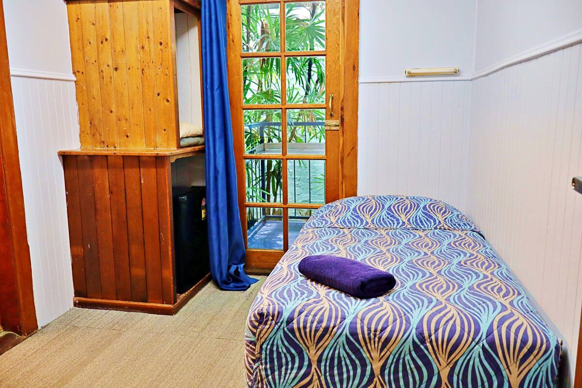 Quarto com cama, escrivaninha de madeira, janela com cortinas e vista para o verde lá fora.