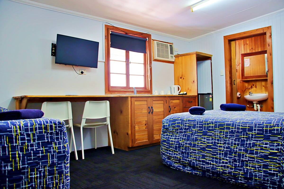 Quarto com duas camas, uma escrivaninha, uma cadeira, uma televisão e uma janela.