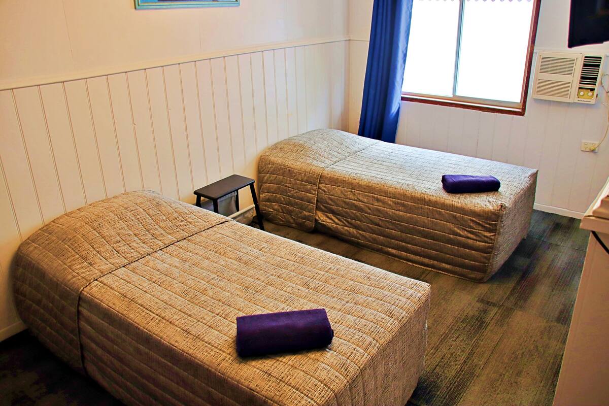 Quarto com duas camas, uma mesinha e uma janela com cortinas.