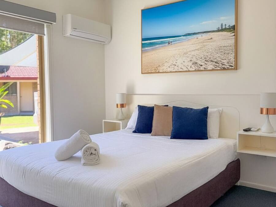 Quarto de hotel com cama, pintura com vista para a praia, ventoinha no teto e janela com unidade de ar condicionado.