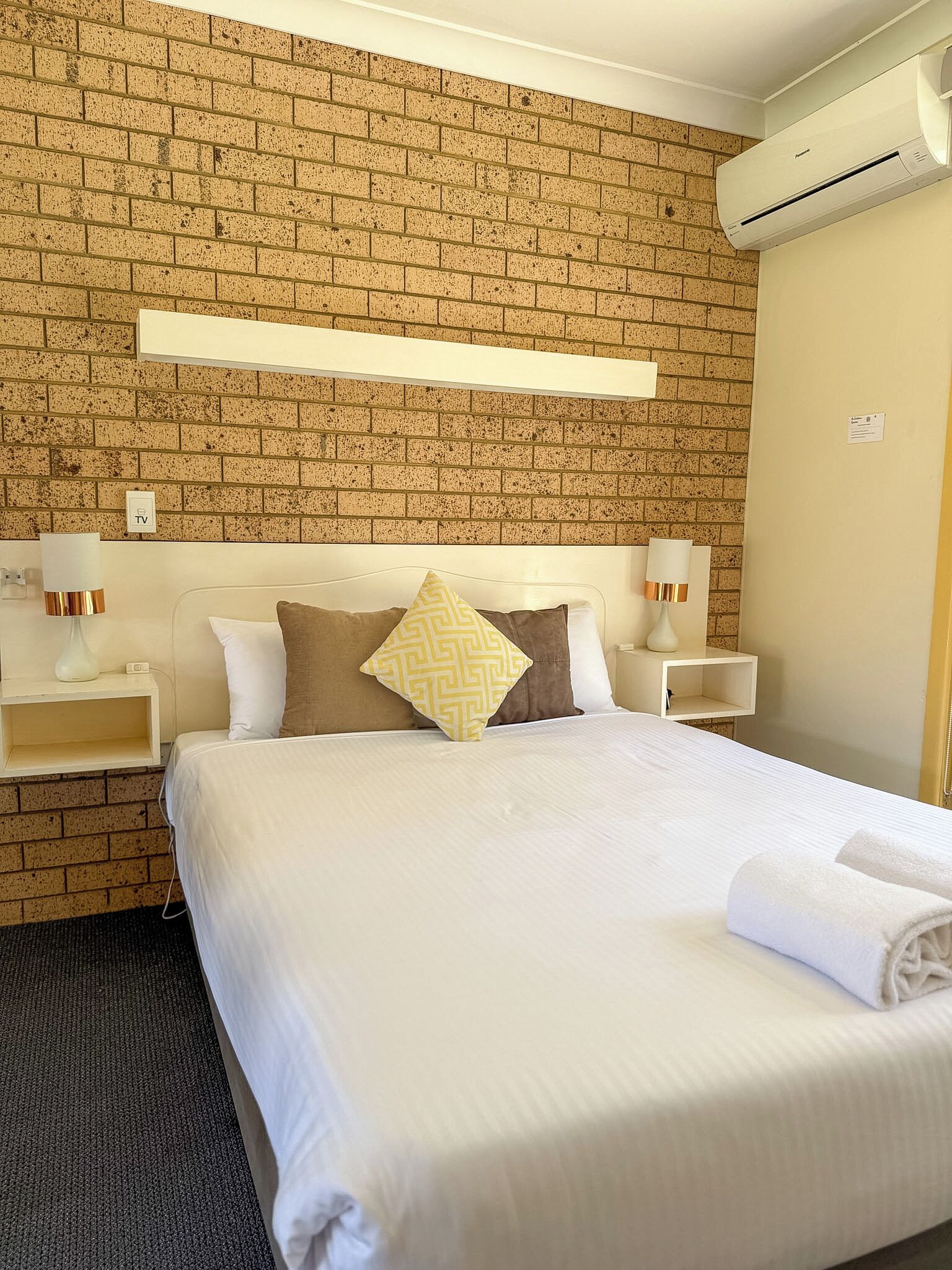 Quarto de hotel com parede em padrão tijolo, cama com roupa de cama branca, duas luminárias de cabeceira e um aparelho de ar condicionado.