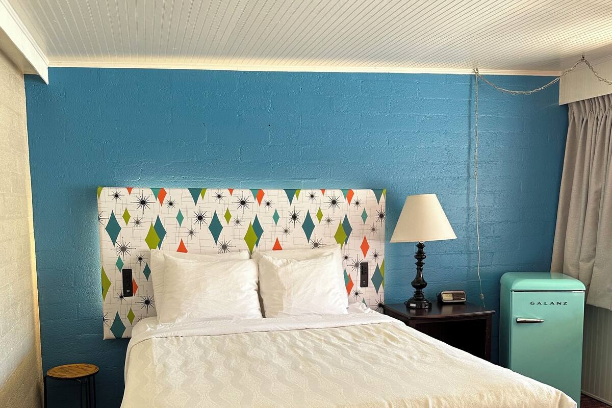 Quarto de hotel com cama, mesa de cabeceira, abajur, pequena mesa e um armário azul.