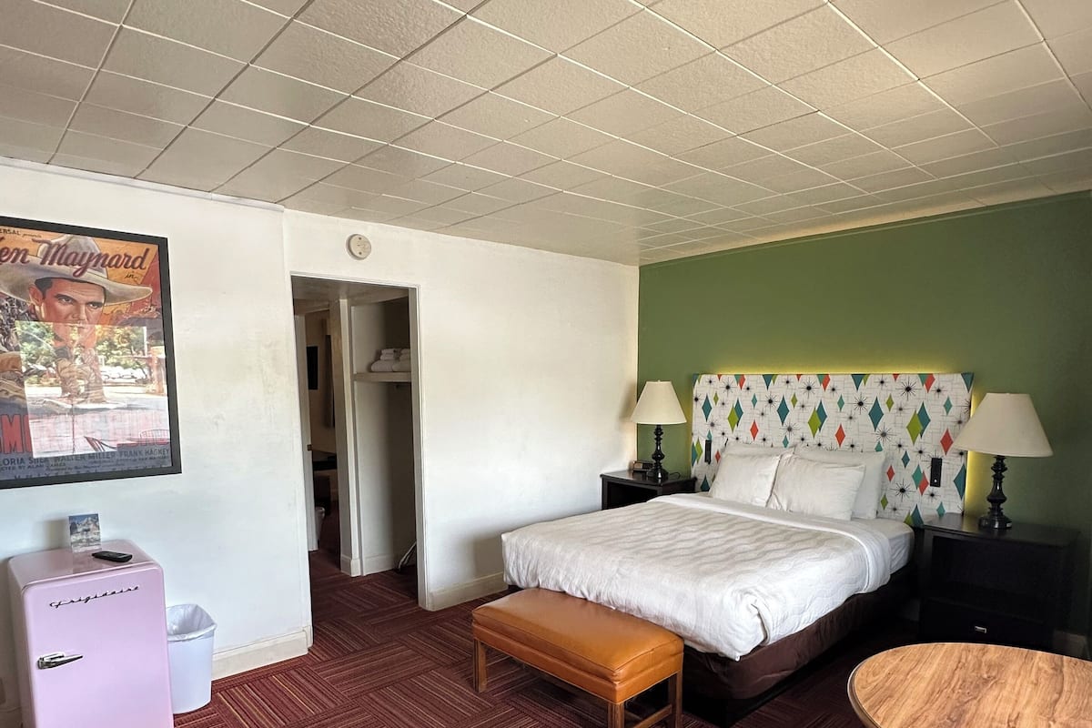 Quarto de hotel com cama, mesas de cabeceira, uma pequena mesa, um banquinho, lixeira, geladeira e um cartaz na parede.
