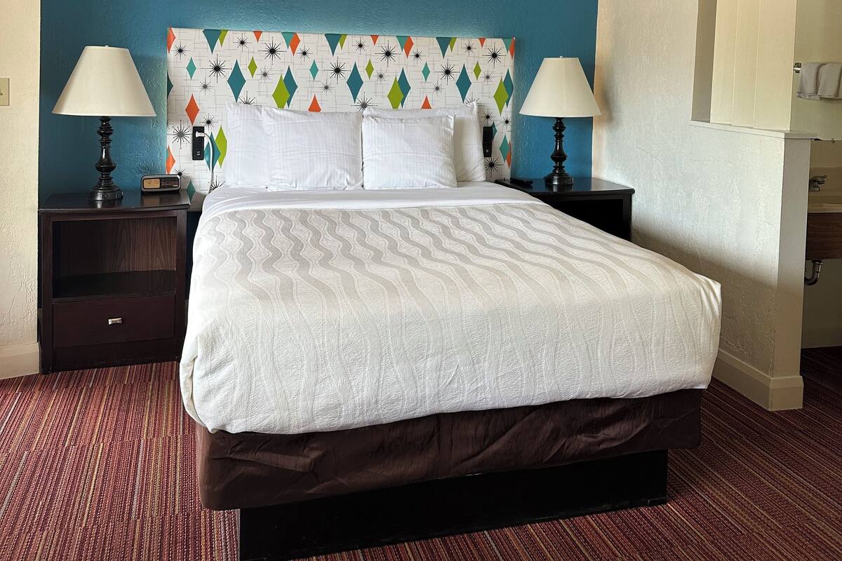 Quarto de hotel com uma cama grande, duas mesas de cabeceira com abajures, uma parede azul com detalhes e uma cabeceira branca com padrão geométrico.