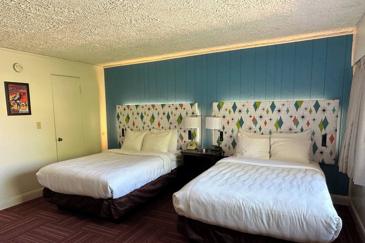 Quarto de hotel com duas camas, cabeceira com padrão geométrico, mesa de cabeceira com luminária e um pôster na parede.