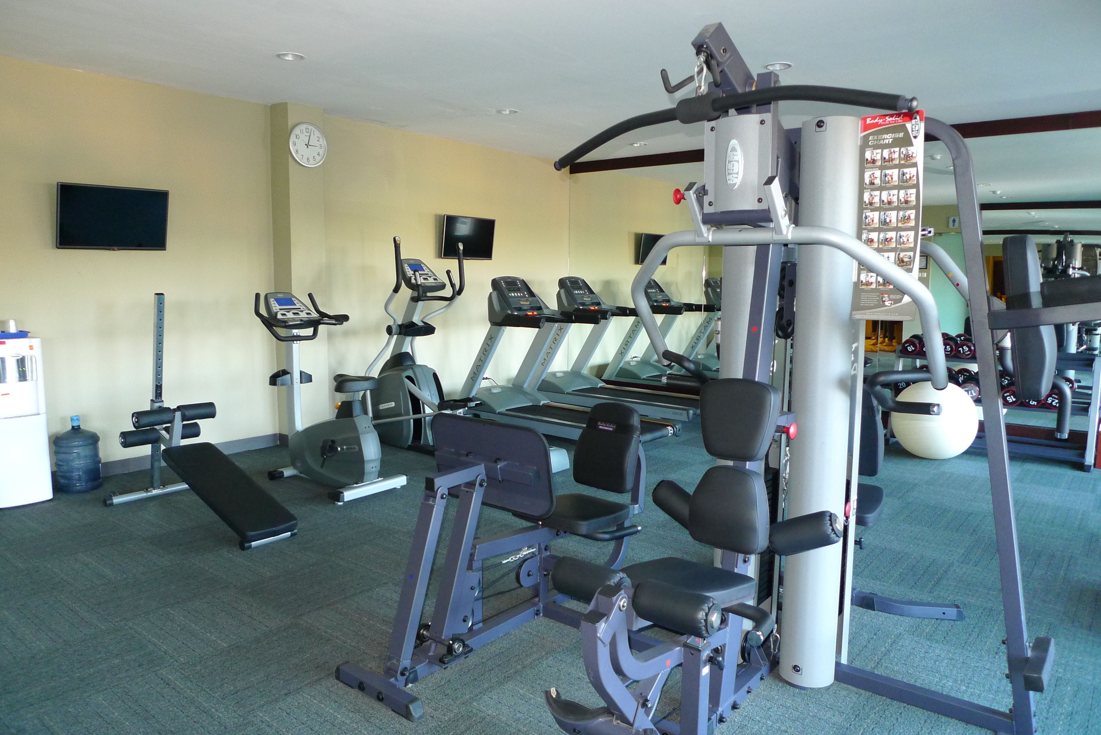 Uma academia com esteiras, máquinas de musculação, aparelho de supino e cooler de água.